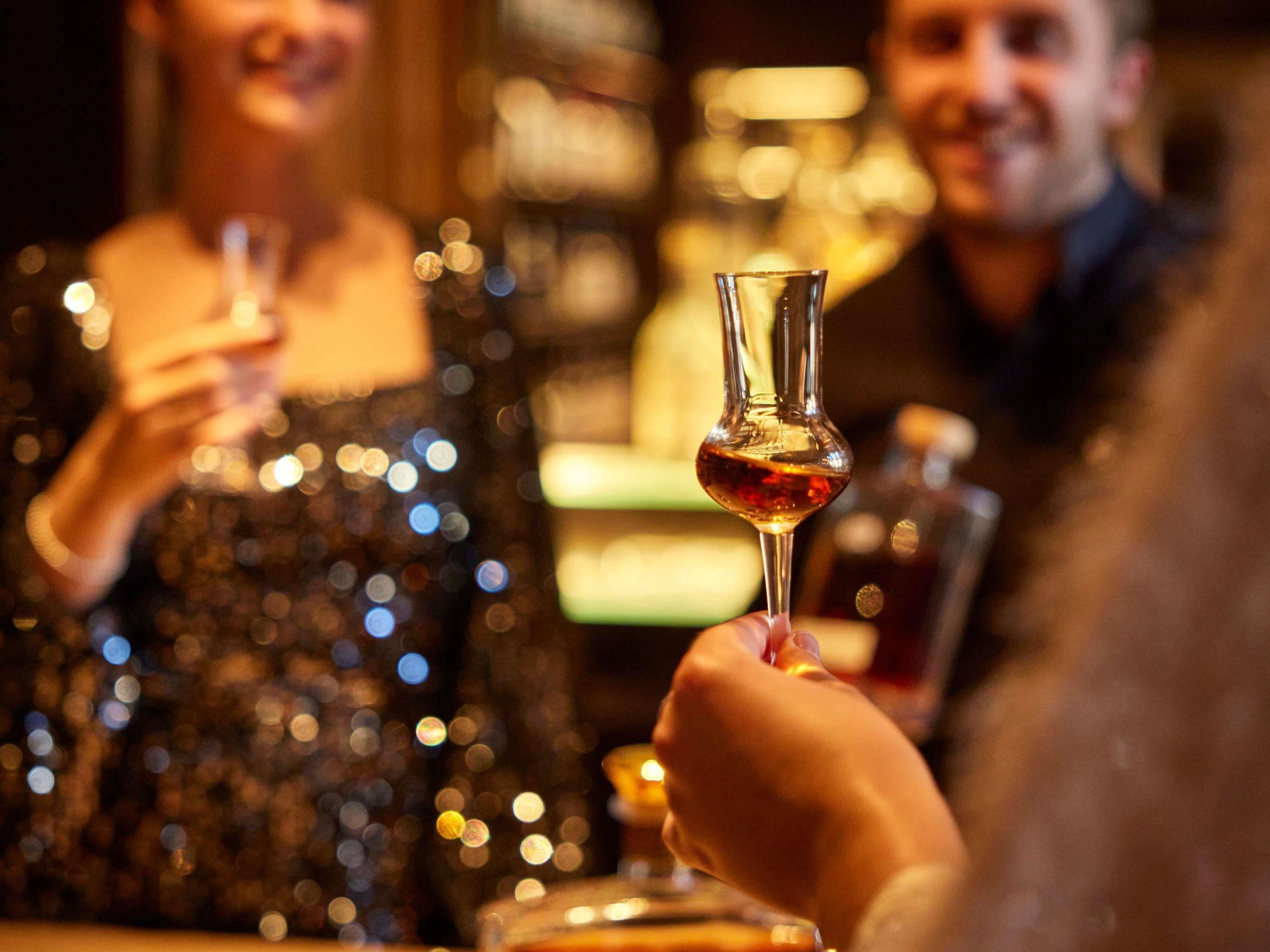 Uma pessoa segurando uma taça de vinho de formato peculiar, em ambiente com pouca luz e luzes desfocadas ao fundo.
