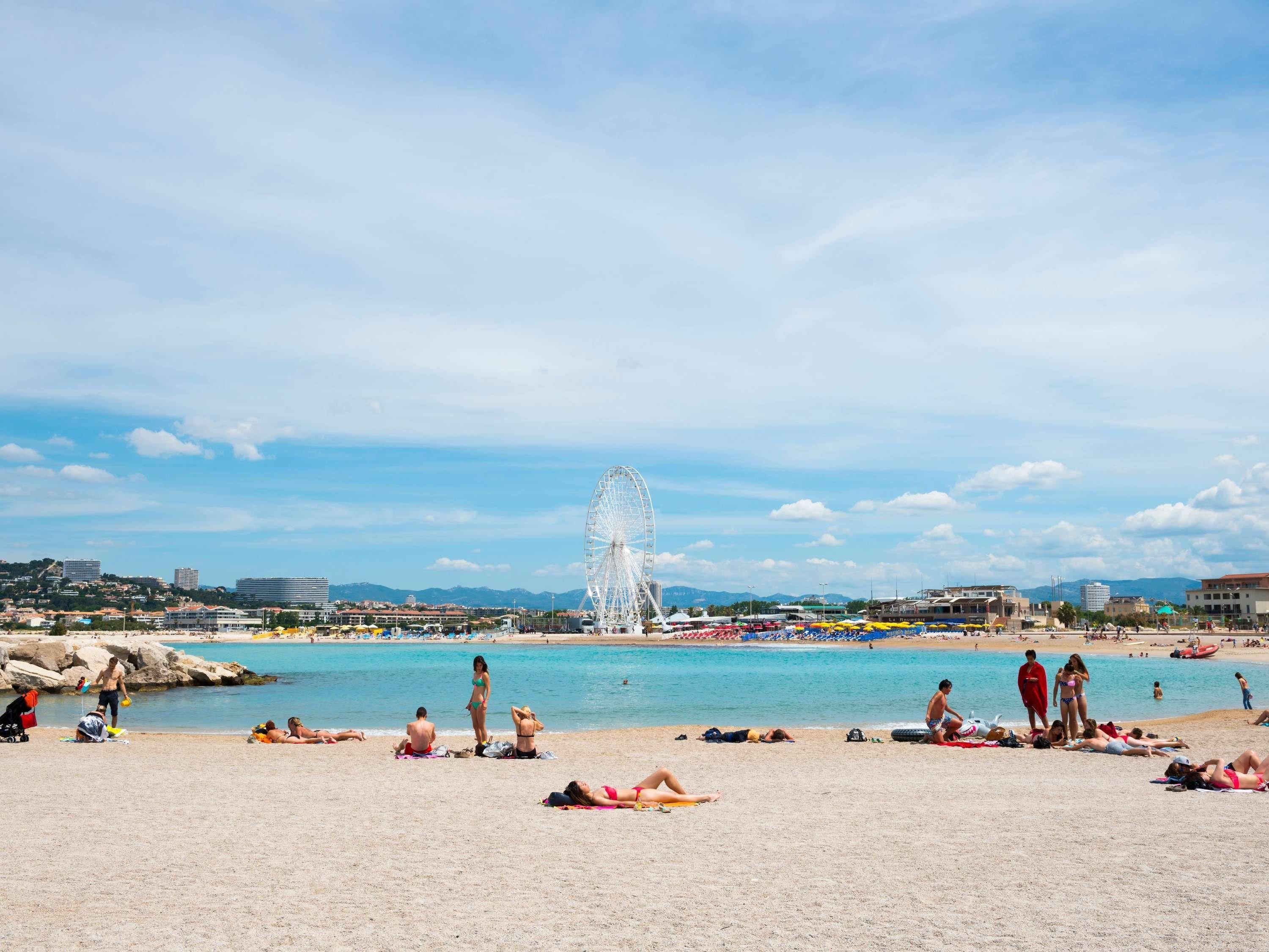 Uma praia com pessoas se banhando e tomando sol, uma roda-gigante ao fundo e céu claro.
