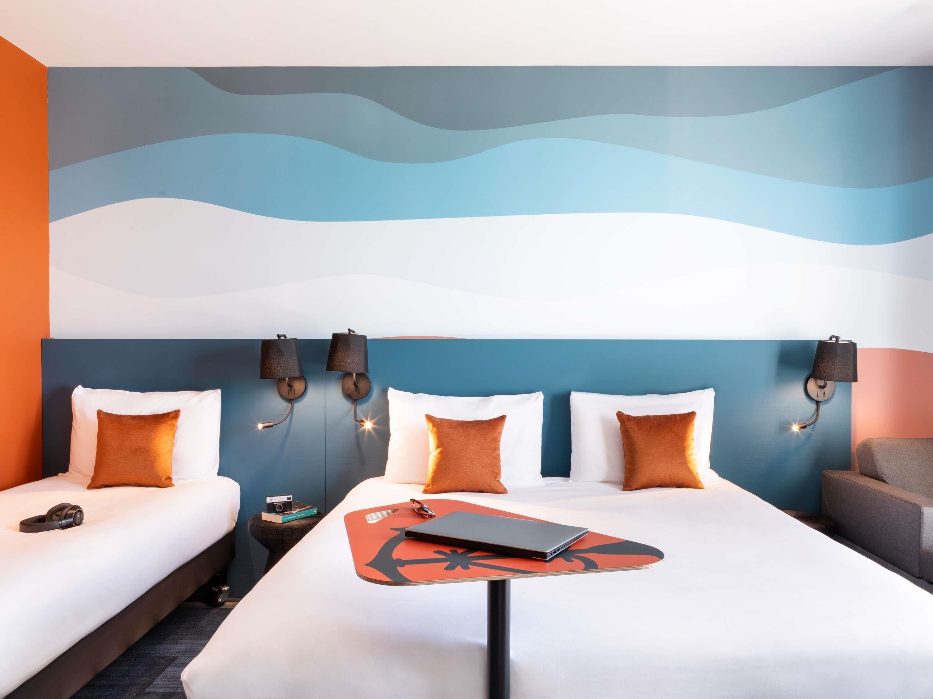 Quarto de hotel com uma cama, duas mesas de cabeceira, uma escrivaninha com um laptop e luminárias fixadas na parede.