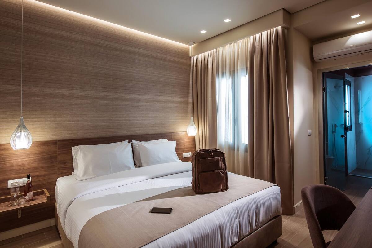 Quarto de hotel com cama, mesa de cabeceira, luminária, cortinas e ar condicionado.