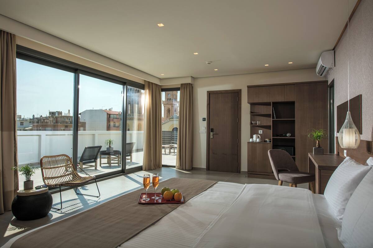 Quarto de hotel moderno com uma cama grande, varanda com assentos e vista para a paisagem urbana.