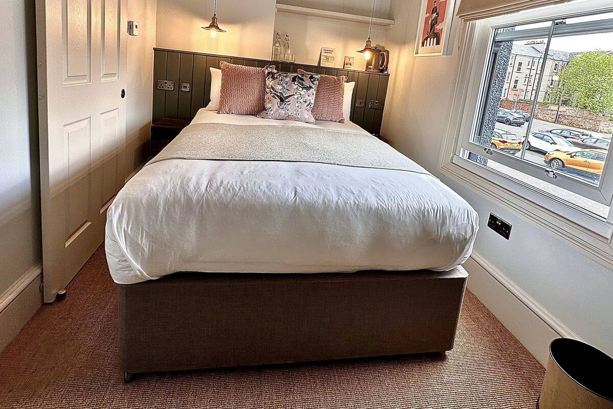 Um quarto com uma cama grande, uma cômoda, uma janela vista de carros e uma luminária no teto.