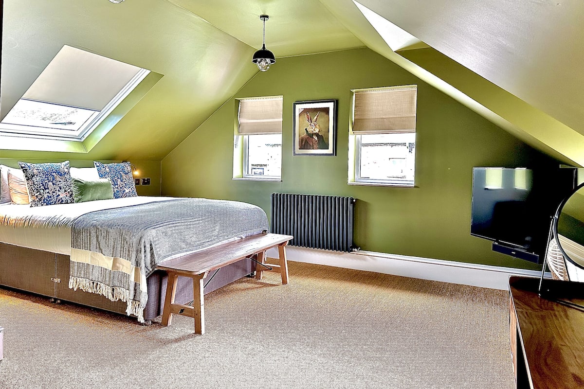 Quarto com teto inclinado, cama com roupa de cama estampada, um banco, duas janelas com persianas, radiador e televisão.