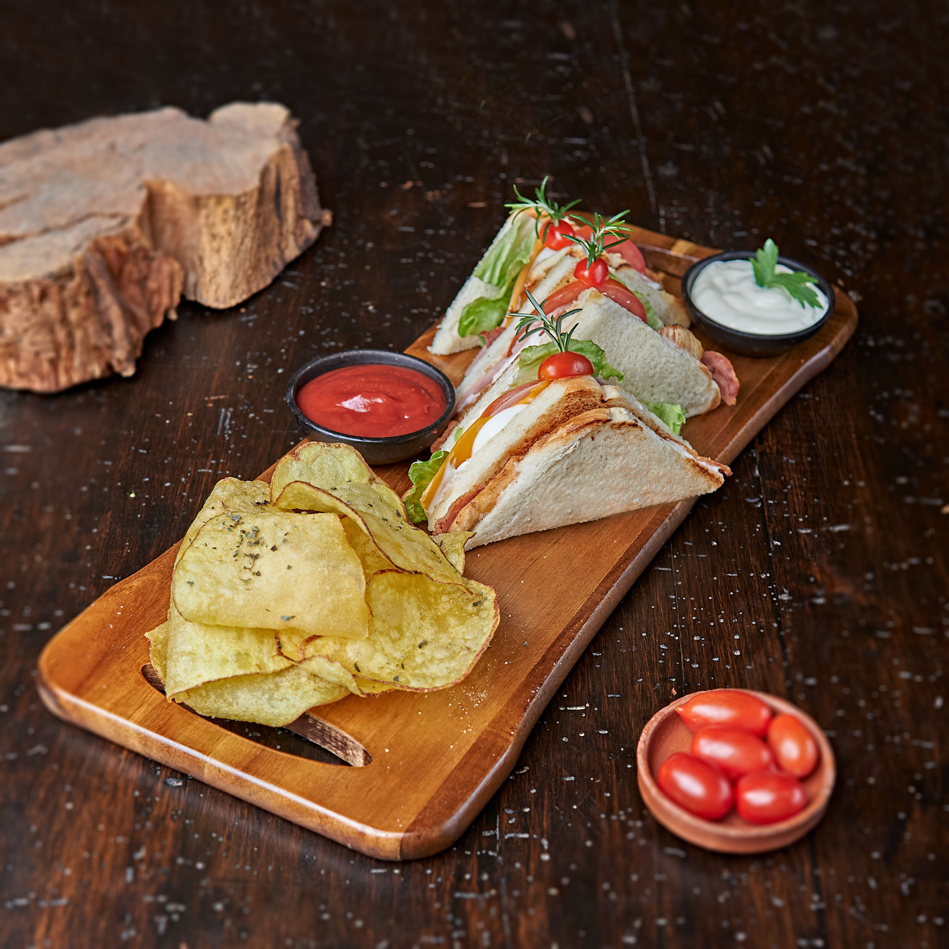 Uma tábua de madeira com sanduíche, batatas fritas e pequenos recipientes de acompanhamentos.