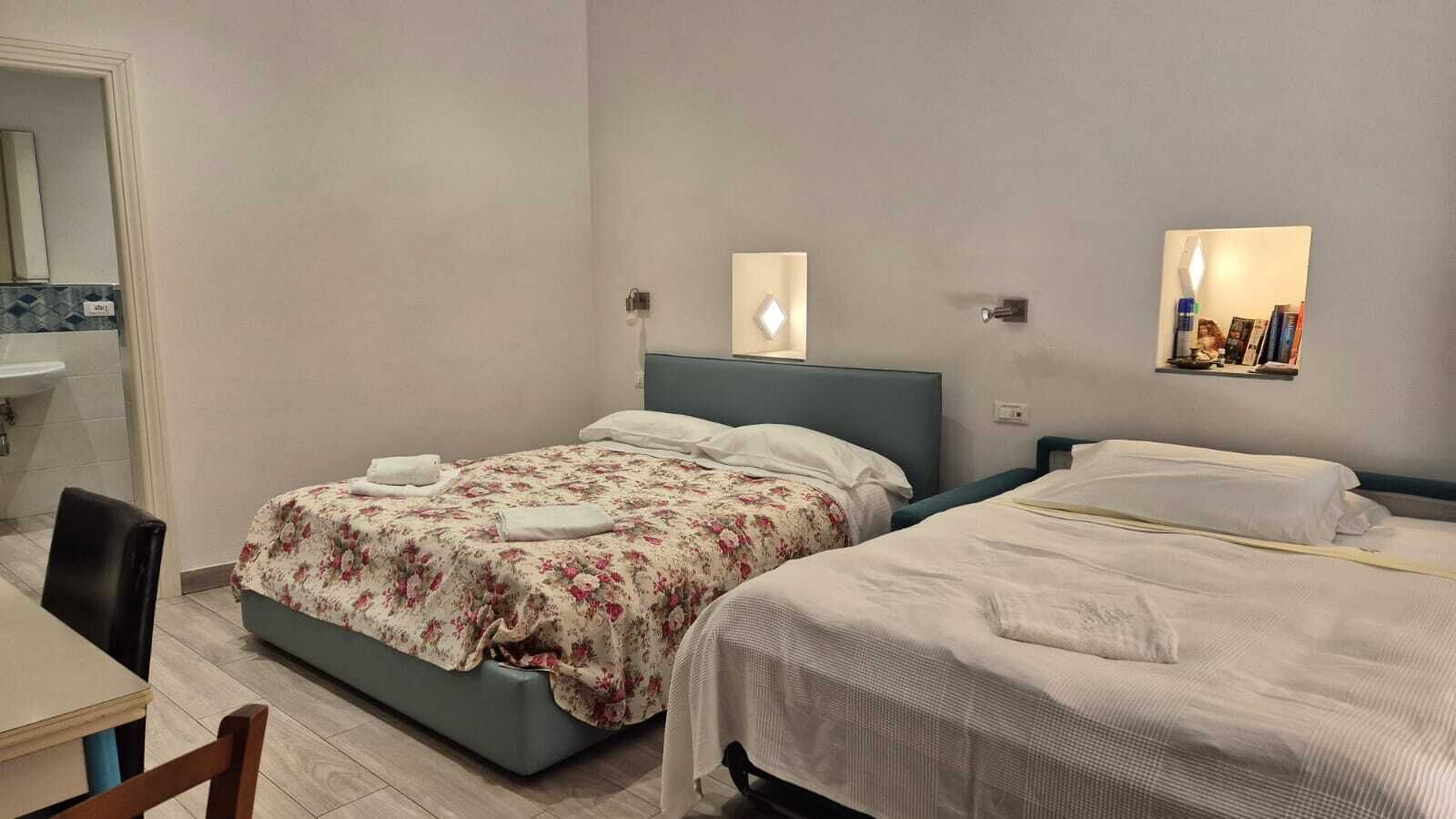 Quarto de hotel com duas camas, uma mesinha, uma cadeira e um espelho.
