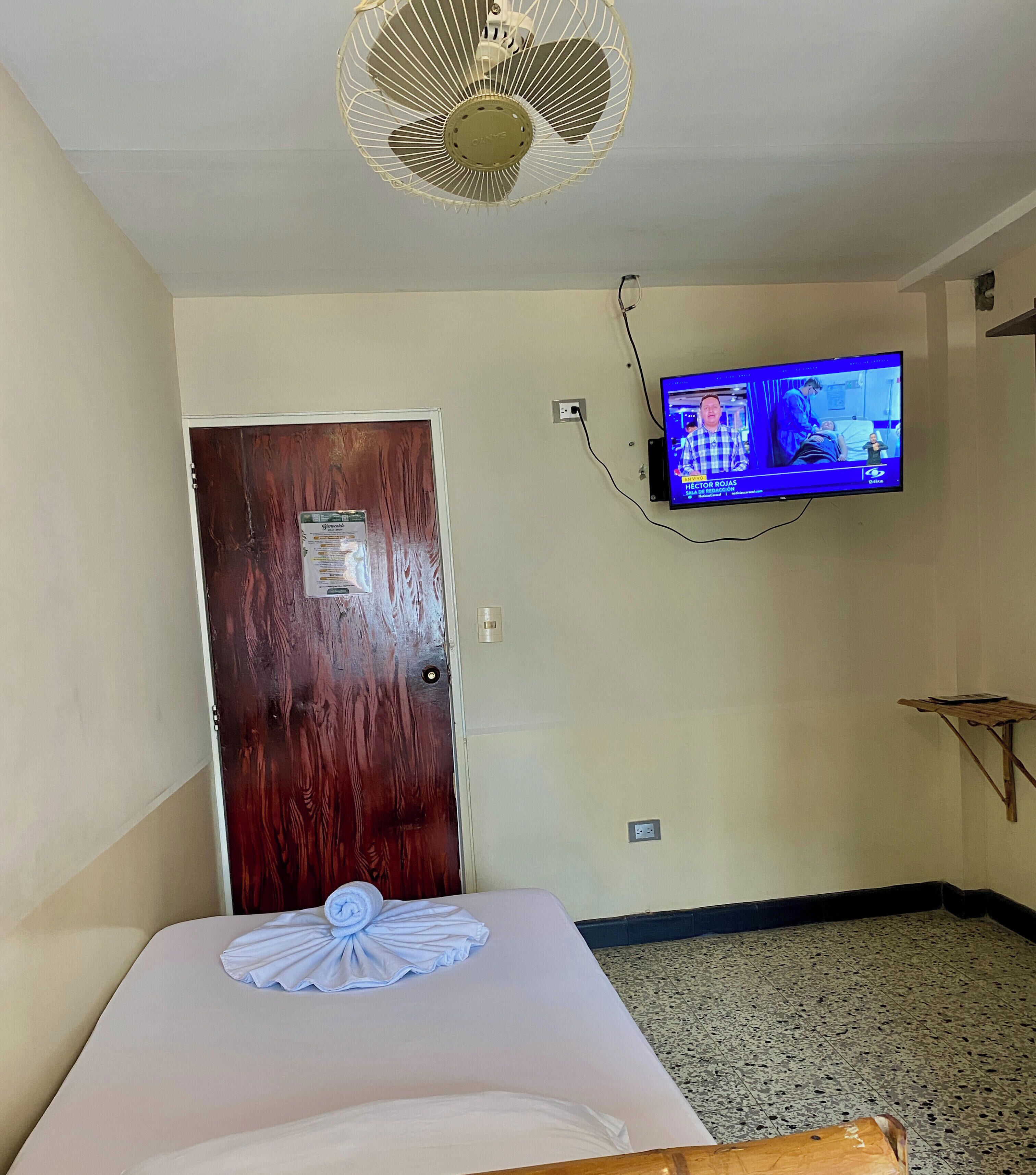 Quarto de hotel com cama, porta de madeira, televisão fixada na parede e ventoinha no teto.