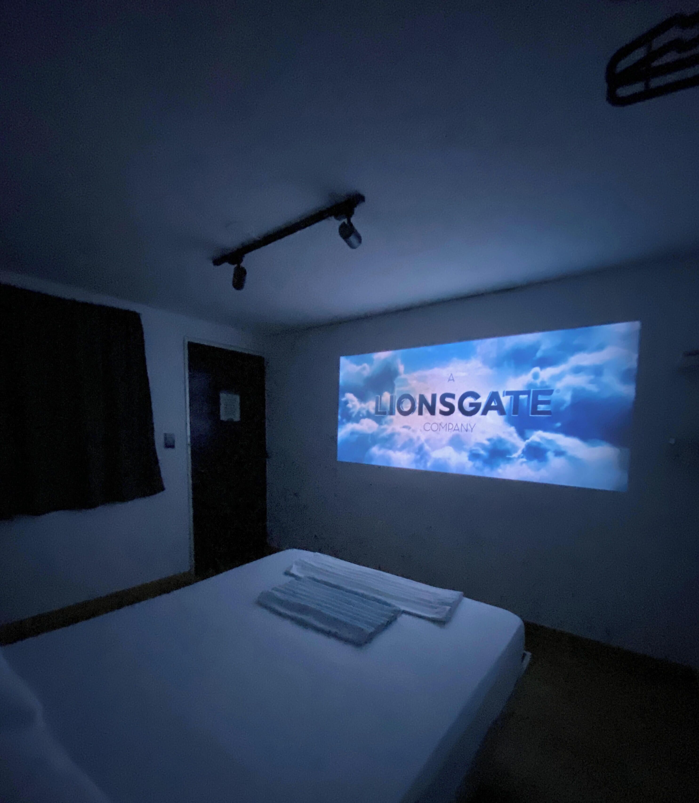 Quarto de hotel com cama, tela de projetor exibindo 'LIONSGATE COMPANY' e luminária fixada no teto.