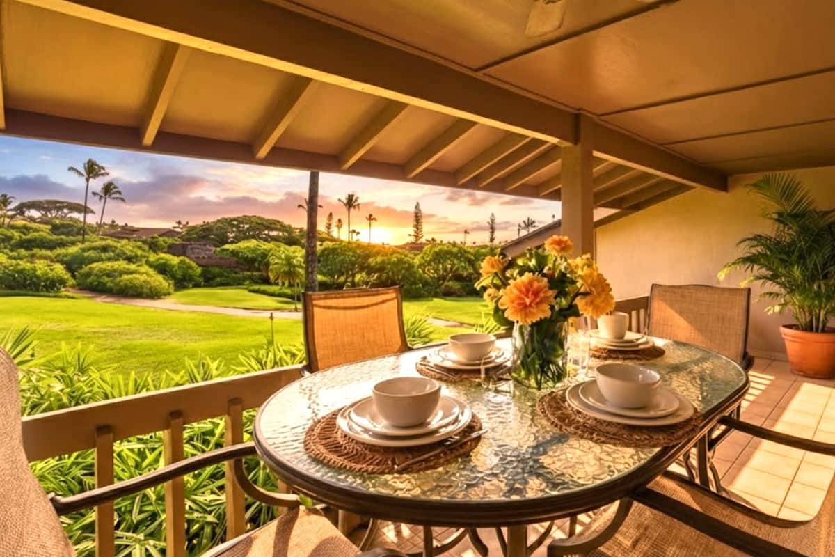 Mesa de vidro redonda com pratos brancos e um vaso de flores, colocada numa varanda com vista para um campo de golfe e o pôr do sol.