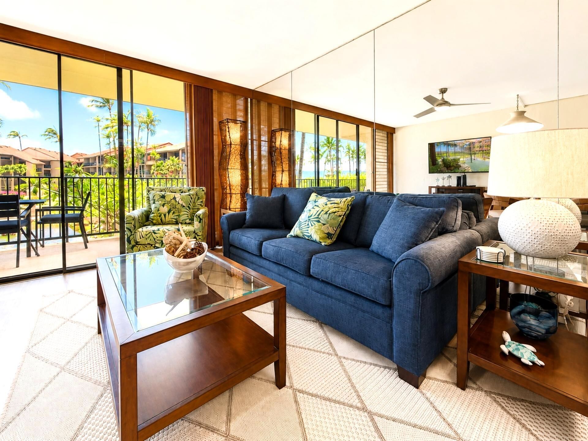 Sala de estar moderna com sofá azul, mesa de centro de vidro e vista para uma paisagem tropical através de grandes janelas.