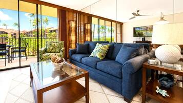 Sala de estar moderna com sofá azul, mesa de centro de vidro e vista para uma paisagem tropical através de grandes janelas.