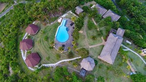 Vista aérea de uma propriedade com piscina, várias estruturas com telhados de palha e vegetação exuberante.