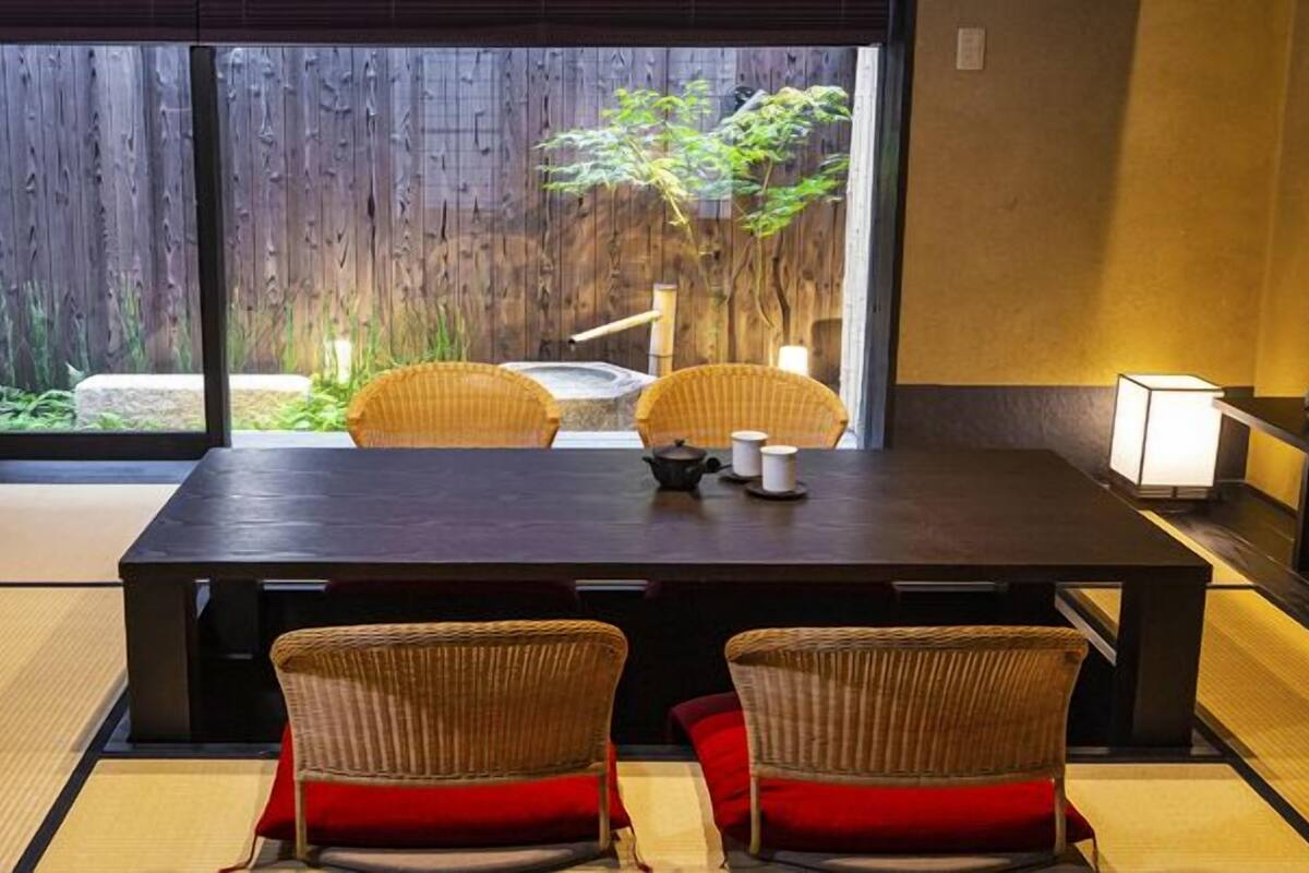 Um quarto em estilo japonês tradicional com tatames, uma mesa baixa, duas cadeiras de vime e vista para uma cerca de madeira e vegetação lá fora.