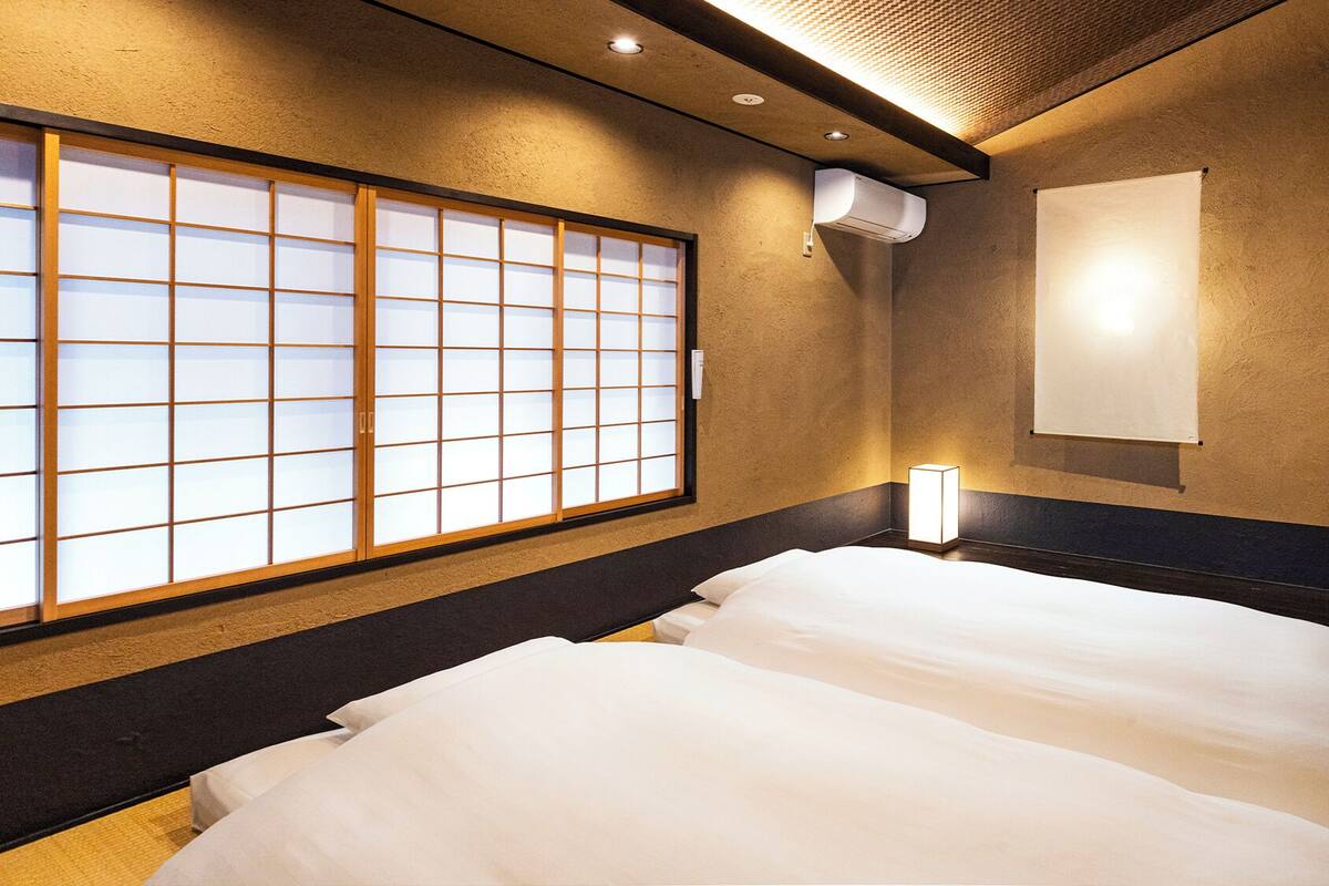 Quarto de hotel com tradicional tela japonesa shoji, cama com roupa de cama branca, ar-condicionado fixado na parede e abajur.