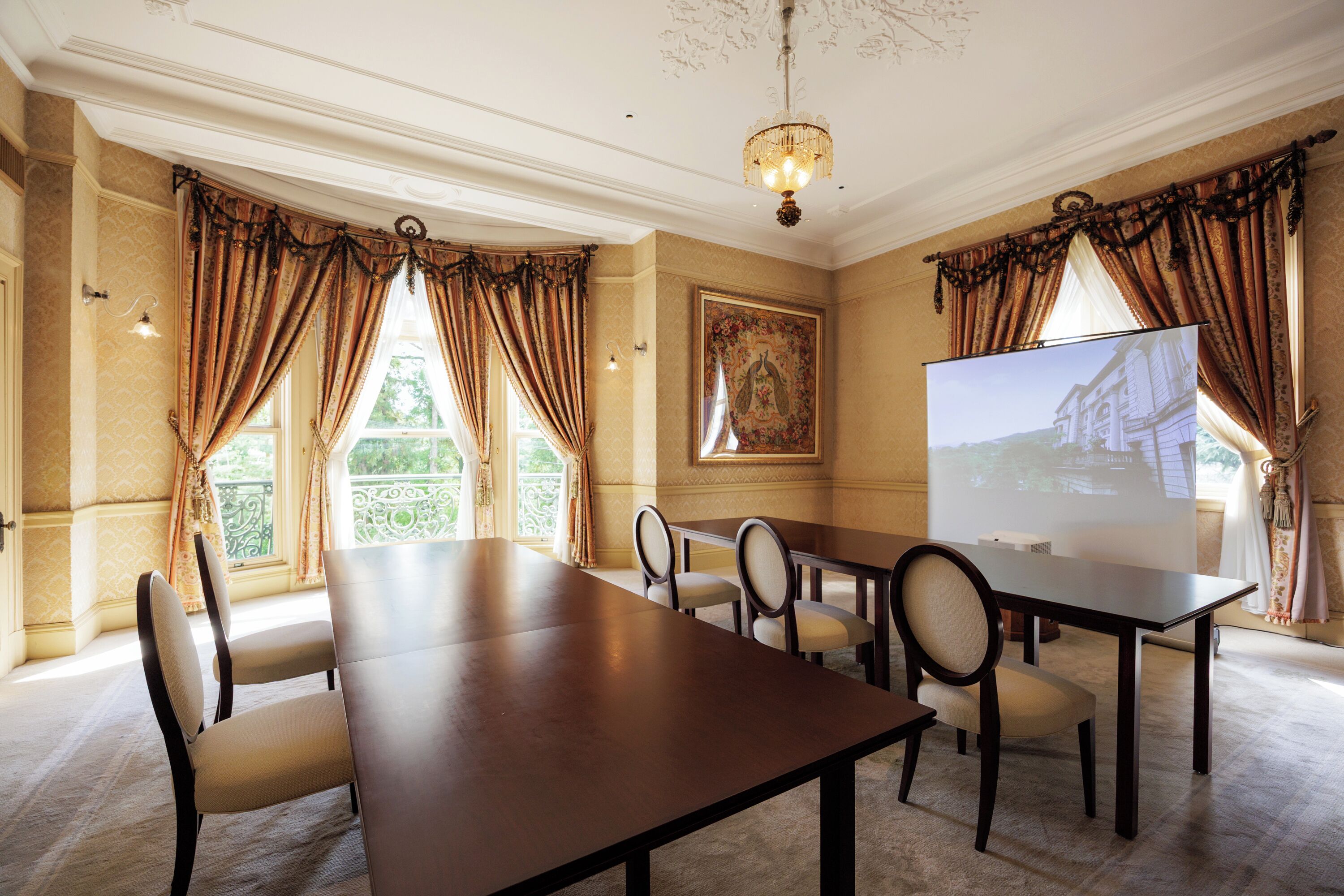 Uma sala de conferências com uma mesa comprida, cadeiras, tela de projetor e grandes janelas com cortinas.