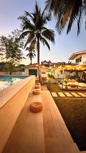 Uma área à beira da piscina com um deck de madeira, uma palmeira e um espaço de estar com almofadas e uma pequena mesa.