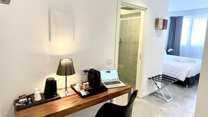 Quarto de hotel com escrivaninha de madeira, duas cadeiras pretas, um laptop, uma cafeteira e uma cama com edredom branco.
