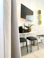 Quarto moderno com televisão de tela plana na parede, uma mesinha com uma planta, duas cadeiras pretas e um espaço compacto para passar roupa.