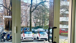 Visão através de uma porta de vidro mostrando uma área de estacionamento com vários carros e a rua visível lá fora.