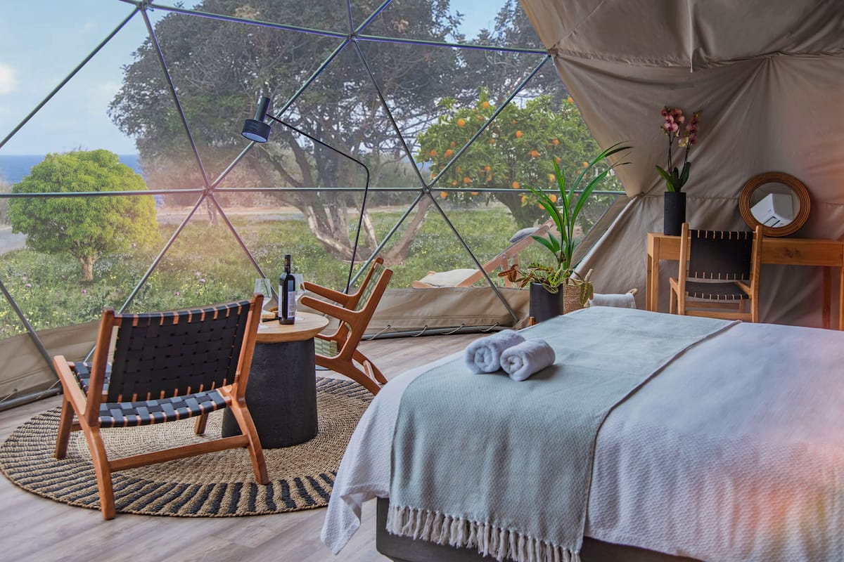 Acomodação em tenda com cama, uma cadeira de madeira, uma mesinha, um espelho e um vaso de planta.