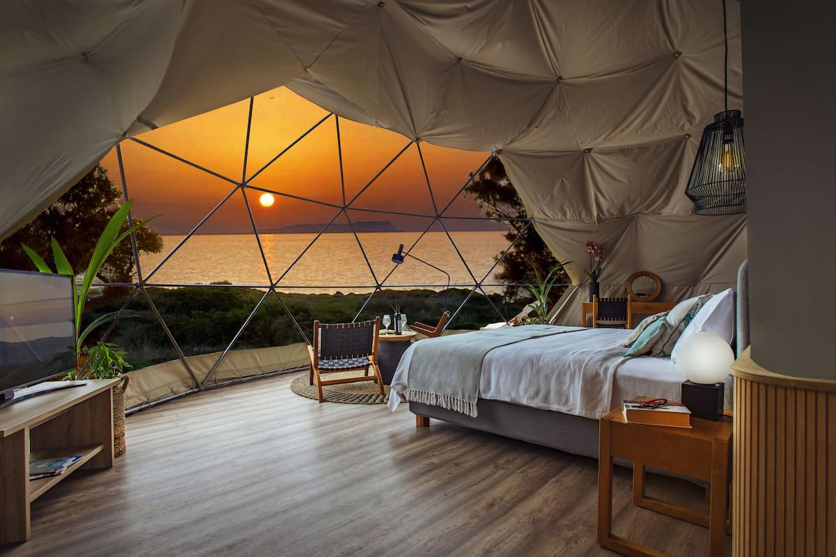 Acomodação em tenda com cama, mesa de cabeceira, luminária e vista para o pôr do sol sobre o mar.