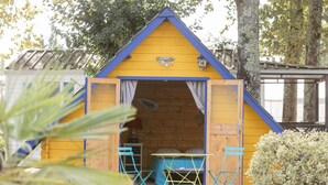 Uma cabana de madeira pequena com exterior amarelo e detalhes em azul, com uma cama, uma mesa e duas cadeiras.