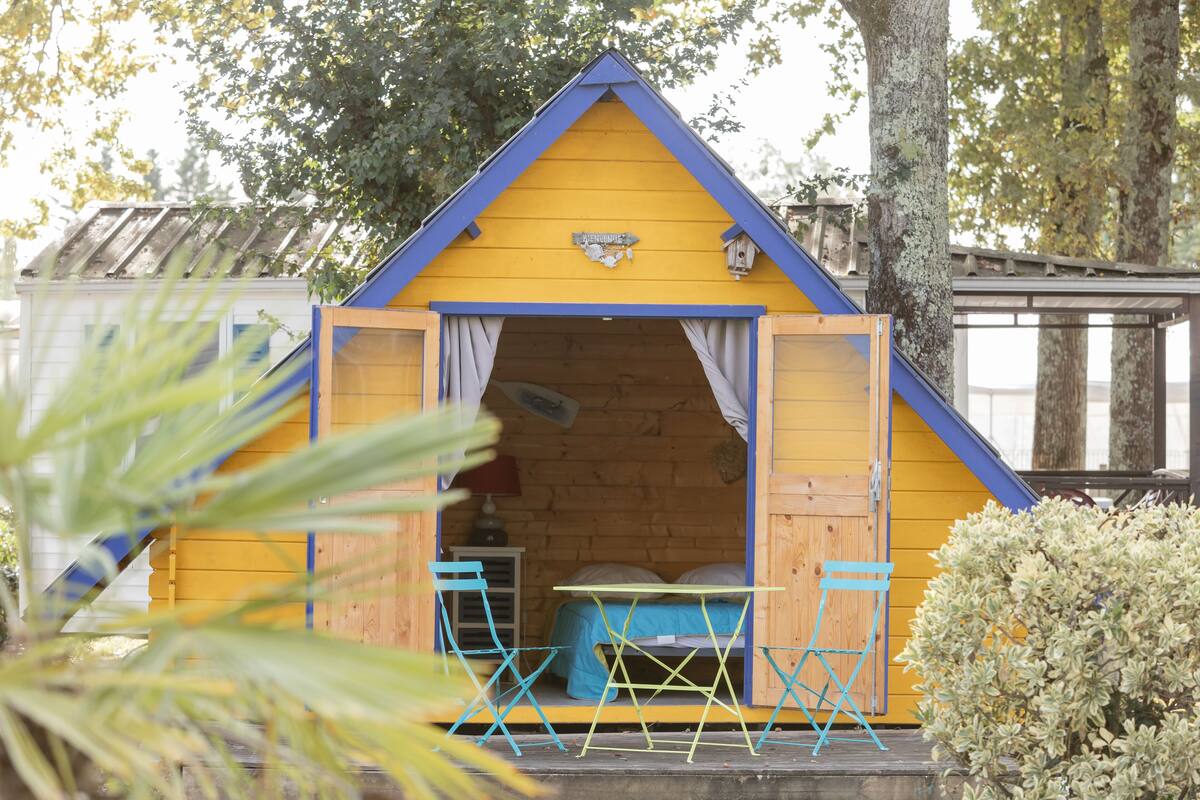 Uma cabana de madeira pequena com exterior amarelo e detalhes em azul, com uma cama, uma mesa e duas cadeiras.
