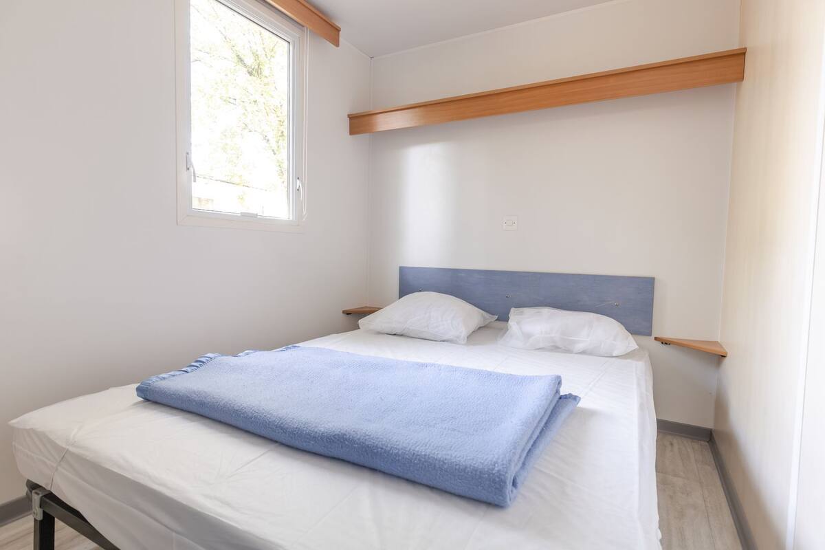 Cama bem arrumada com um cobertor azul, cabeceira de madeira e uma pequena prateleira na parede.