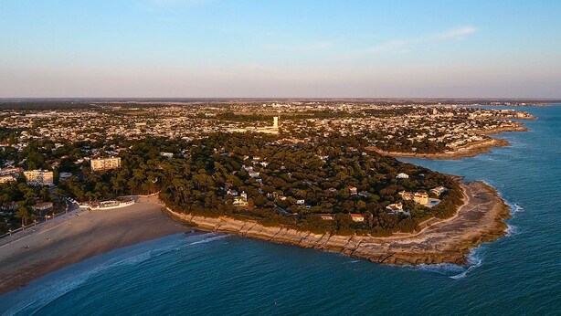Vista aérea de uma vila costeira com um farol imponente, área residencial densa e uma praia.
