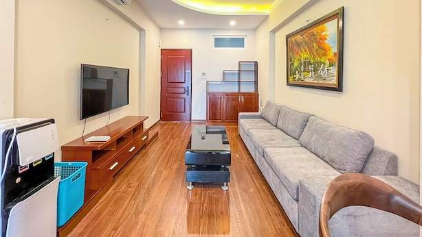 Uma sala de estar moderna com televisão de tela plana, sofá cinza, piso de madeira, teto com iluminação circular e um quadro na parede.