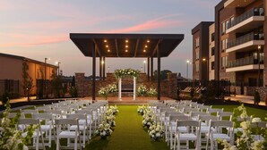 Espaço para casamento ao ar livre, com fileiras de cadeiras brancas, uma passagem central decorada com flores e uma área coberta com iluminação.