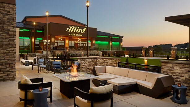 Uma área de estar externa com lareira, paredes de pedra e um edifício com a inscrição "The Mint Event Center" ao fundo.