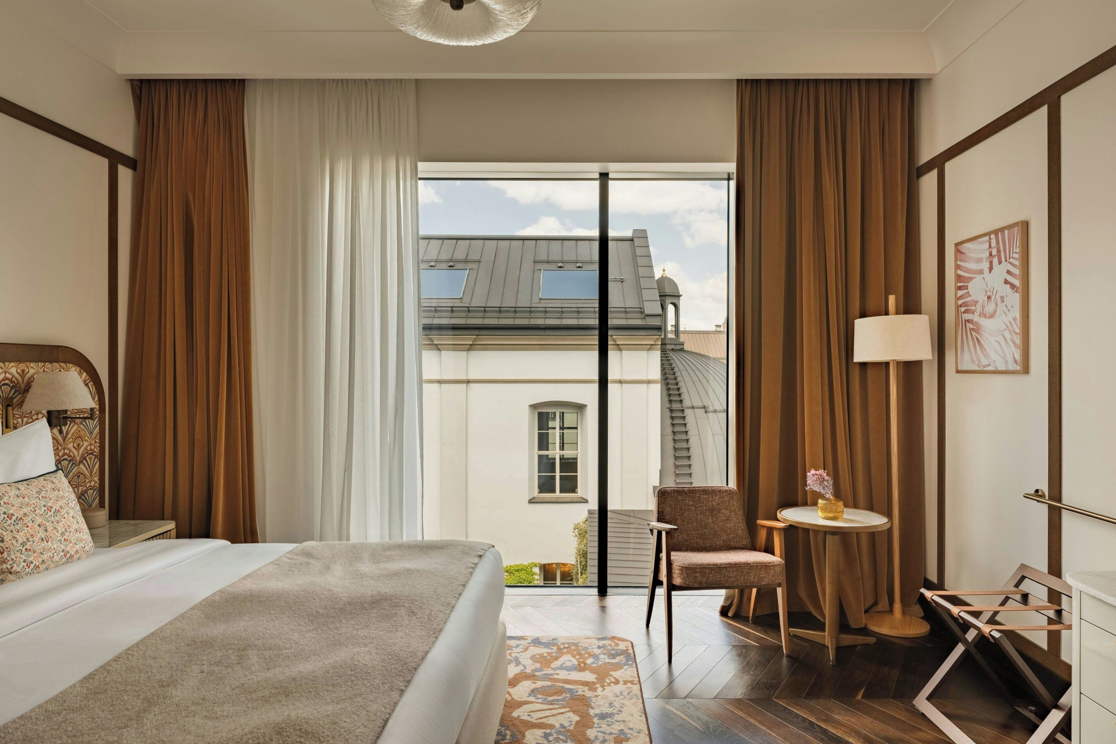 Quarto de hotel com cama, poltrona, mesinha, luminária e vista para o exterior.
