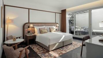 Quarto de hotel com uma cama grande, uma escrivaninha, uma cadeira, uma mesinha e vista para o exterior.