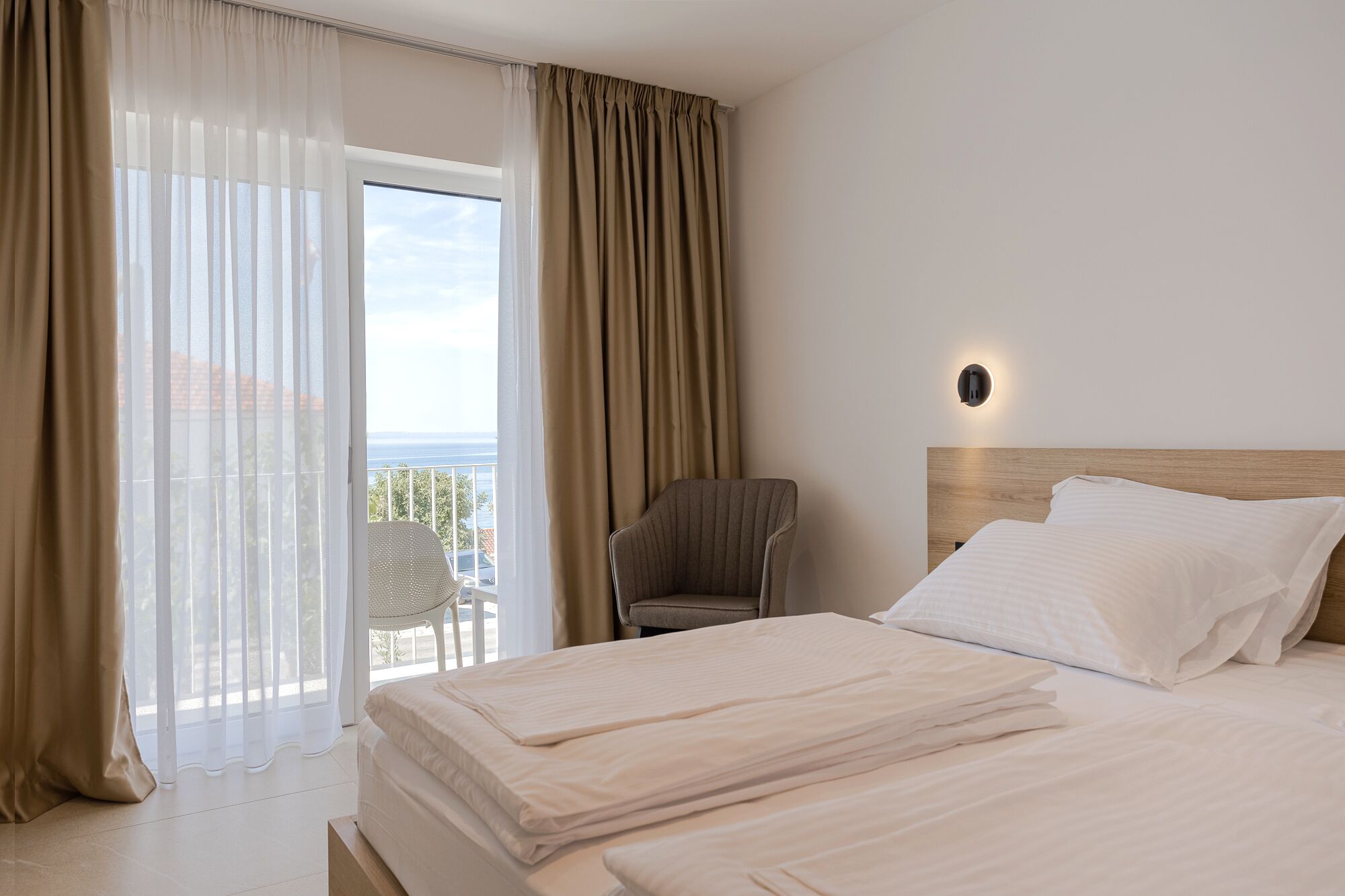 Quarto de hotel com cama, cadeira, varanda e vista, e luminária na parede.