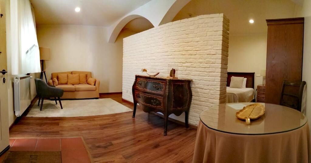 Quarto de hotel com piso de madeira, parede de tijolos brancos, um sofá, uma mesa pequena com um objeto decorativo e uma cama com cabeceira de madeira.