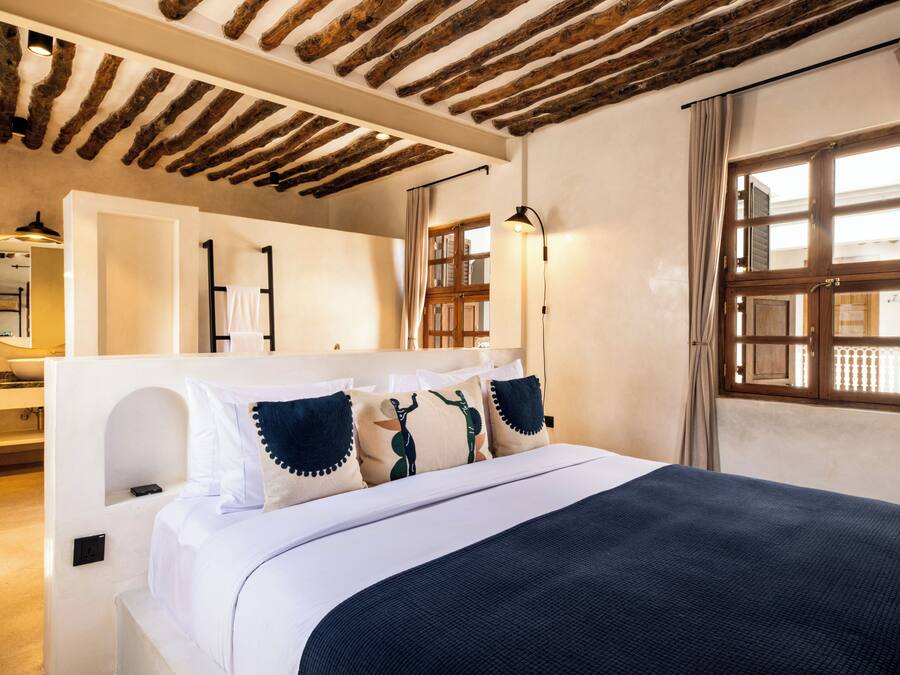 Quarto com cama grande, vigas de madeira no teto, janela com cortinas e um espelho na parede.