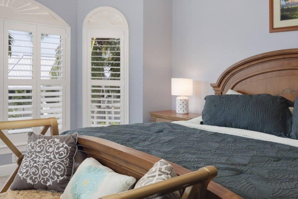 Quarto com cama de madeira, mesa de cabeceira com abajur e duas janelas emolduradas a branco com persianas.
