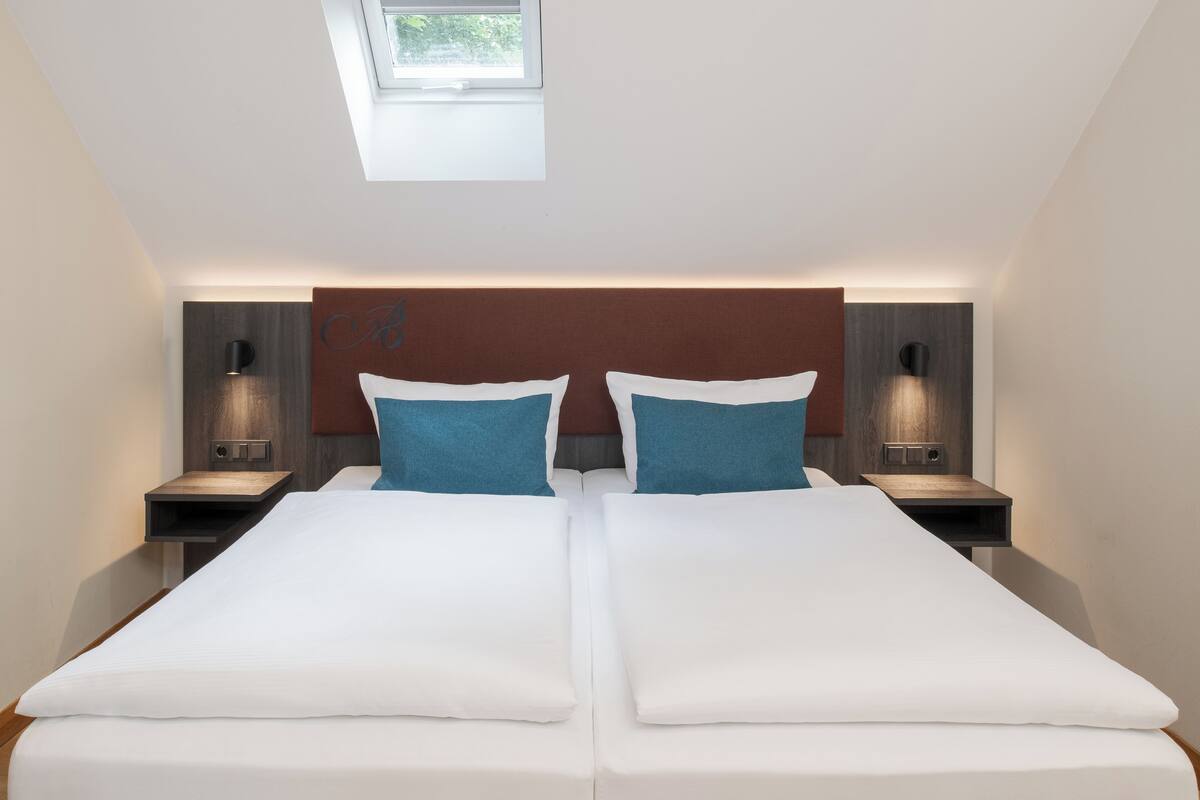 Uma cama de casal com roupa de cama branca e almofadas azuis, duas mesas de cabeceira com abajures, uma cabeceira de madeira e um claraboia acima.