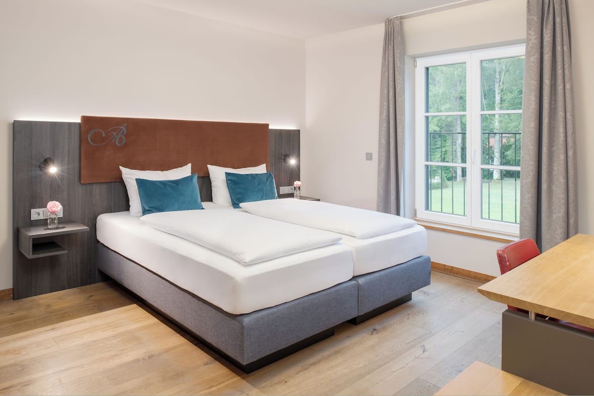 Quarto de hotel moderno com uma cama grande, mesas de cabeceira, escrivaninha de madeira e janela com vista para área verde.