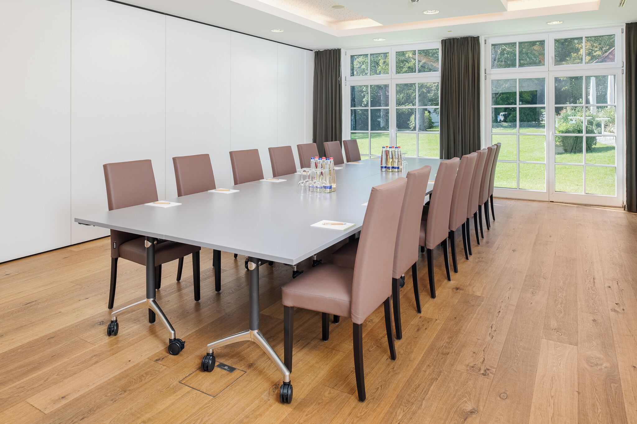 Sala de conferências com uma longa mesa, cadeiras e vista para um jardim através de amplas janelas.