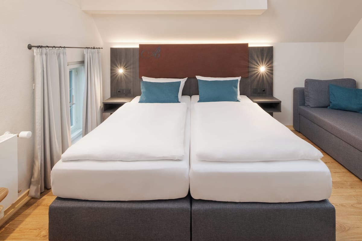 Uma cama de casal com roupa de cama branca e almofadas verde-água, um sofá cinzento e um chão de madeira.