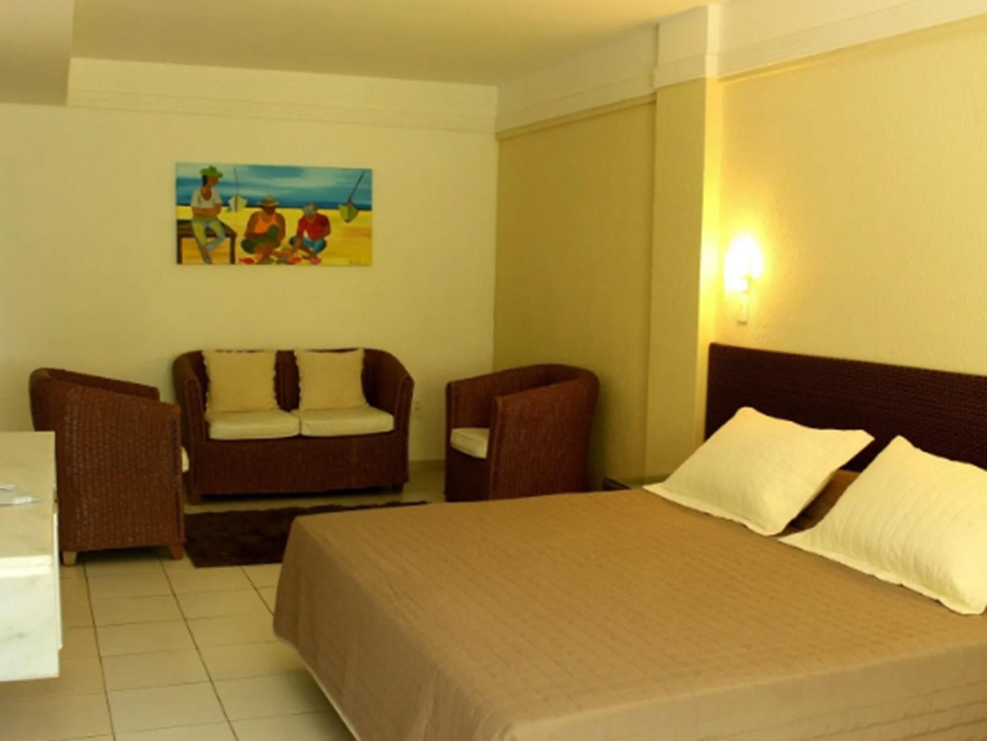 Quarto de hotel com uma cama, um sofá, duas poltronas, um quadro na parede e uma mesinha de cabeceira.