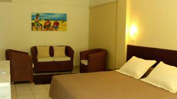 Quarto de hotel com uma cama, um sofá, duas poltronas, um quadro na parede e uma mesinha de cabeceira.