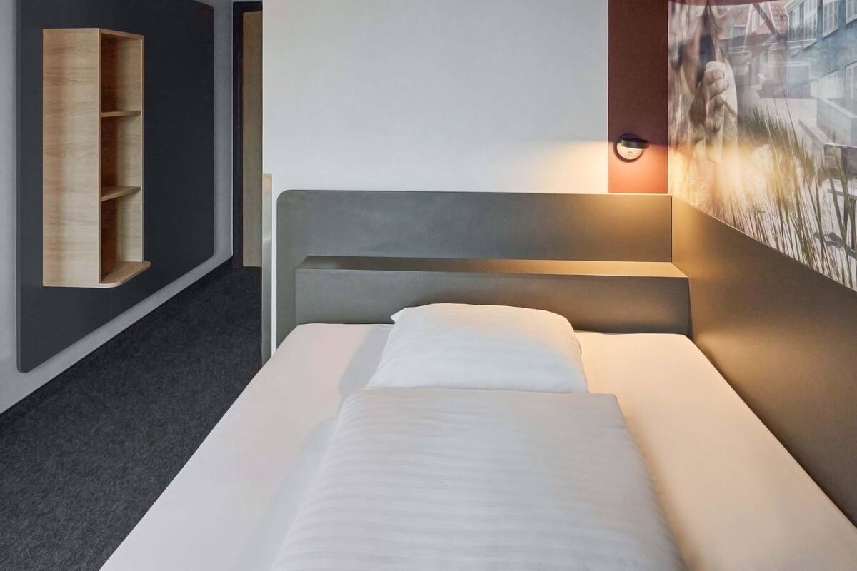Quarto de hotel moderno com cama, estante de madeira e luminária fixada na parede.