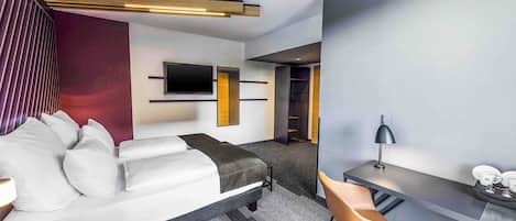 Quarto de hotel moderno com sofá, escrivaninha, cadeira, televisão de tela plana e estante de livros.
