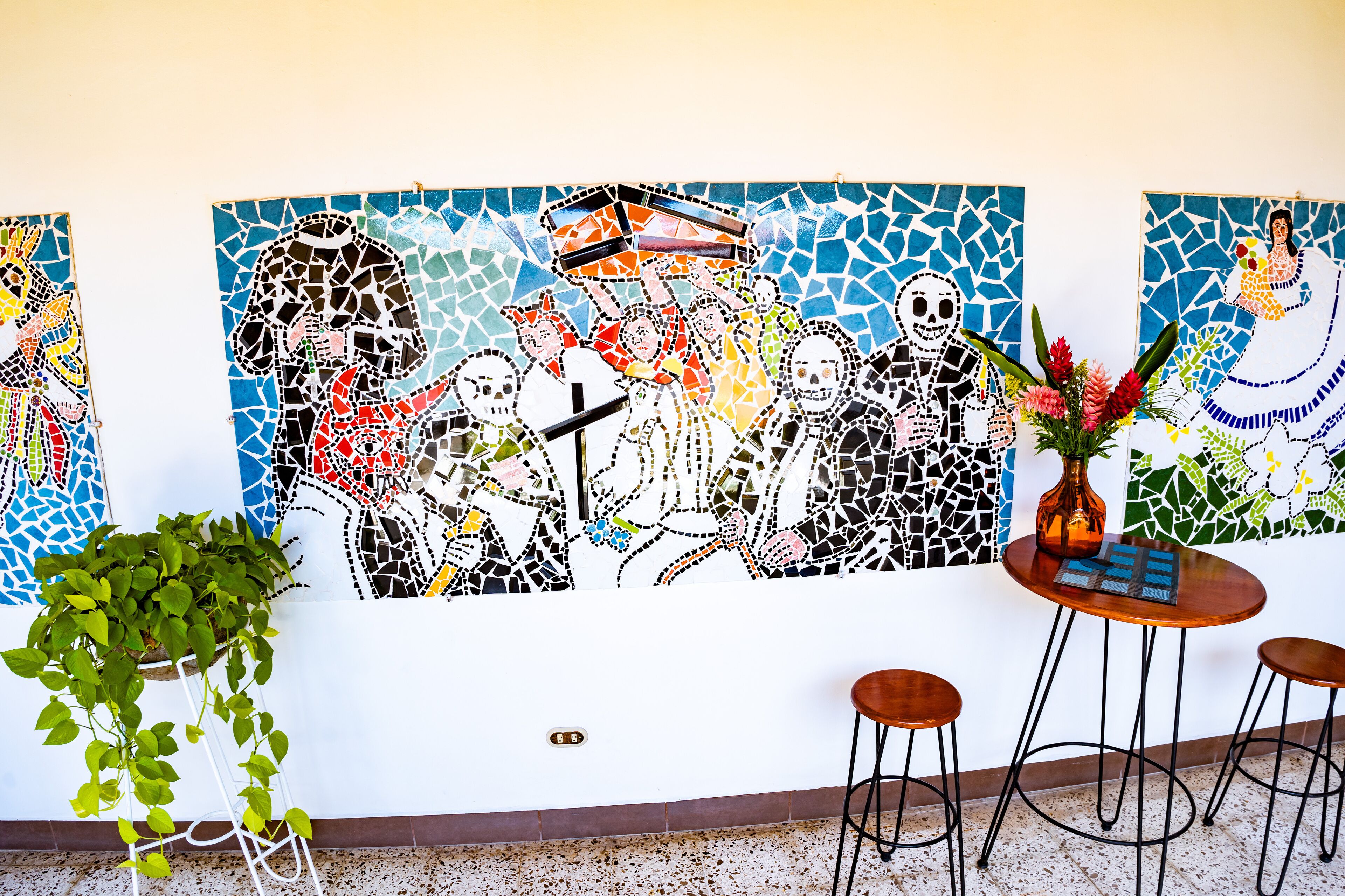Quarto com um grande mural em mosaico, um vaso de planta e uma pequena mesa redonda com bancos.
