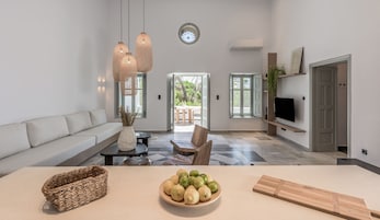 Uma sala de estar moderna com um sofá, uma mesa de jantar e uma TV fixada na parede. Há uma grande janela que oferece luz natural e vista para o exterior.