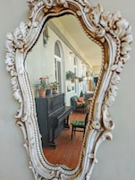 Um quarto com um piano de cauda, uma poltrona estofada em verde e um espelho refletindo um corredor com janelas arqueadas e plantas em vasos.