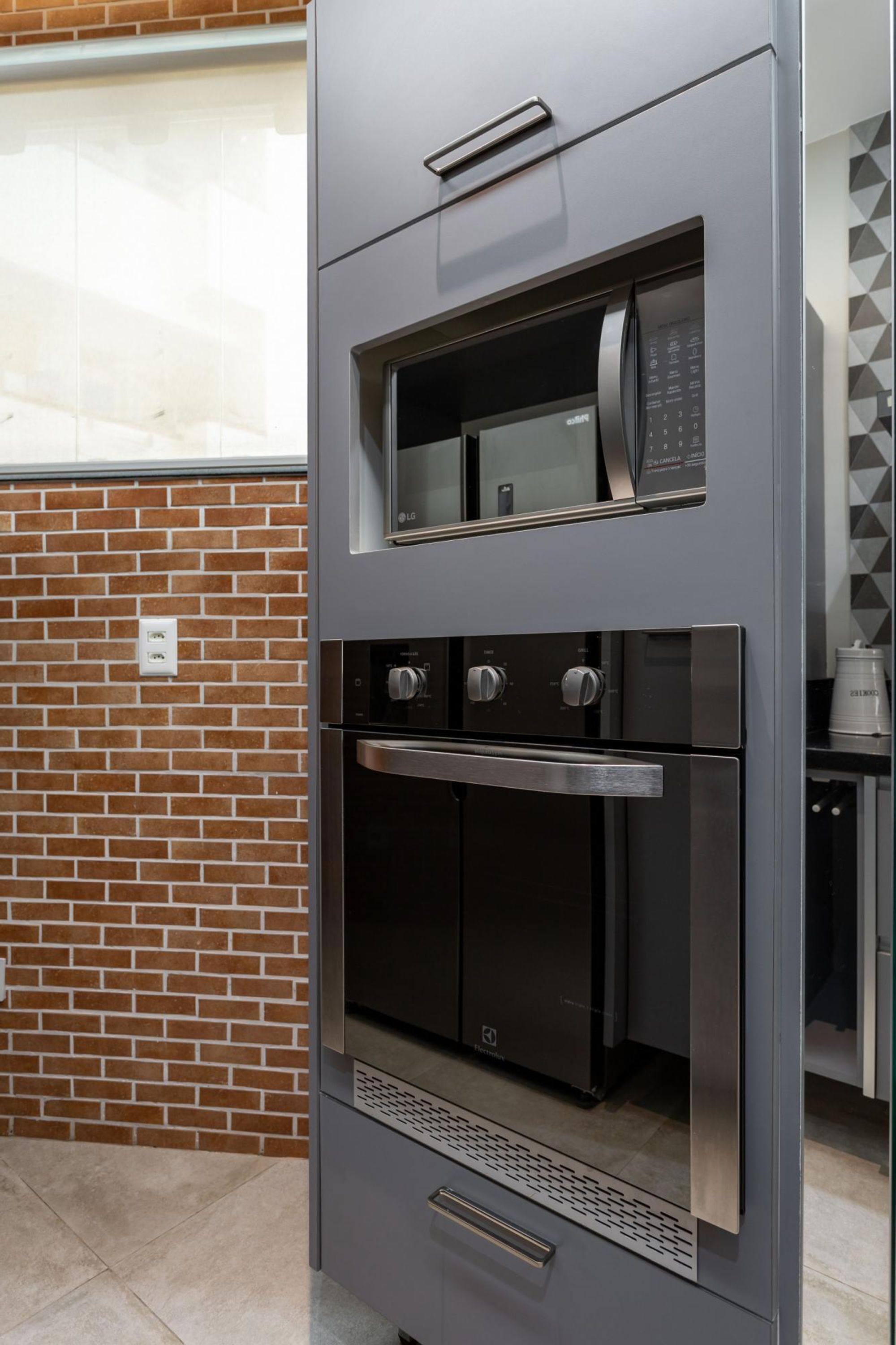 Uma cozinha moderna com forno embutido e micro-ondas, parede de tijolos e chão ladrilhado.