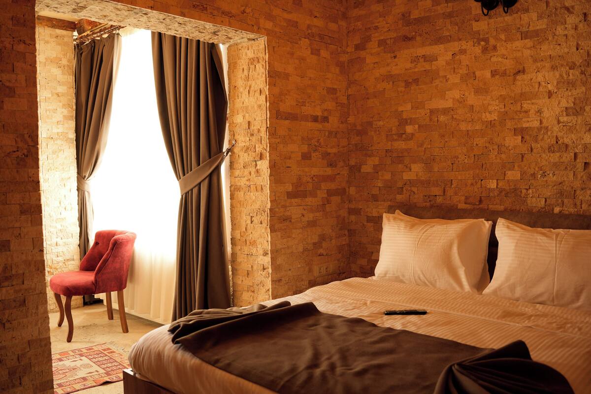 Quarto com parede de tijolos, cama com almofadas brancas, cadeira vermelha e janela com cortinas.